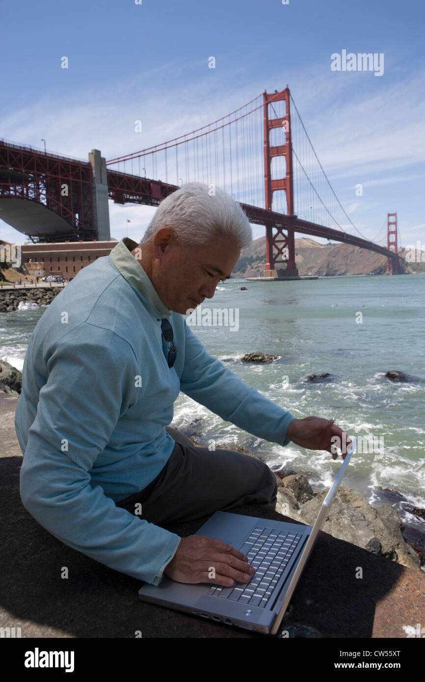 Mature man using a laptop with a suspension bridge in the background ...