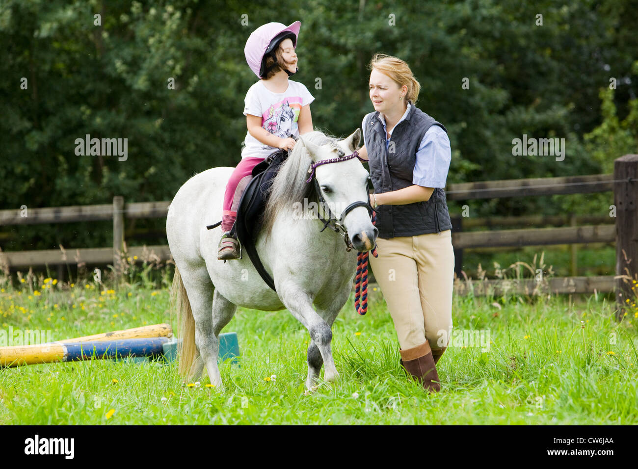 A young girl on a white pony being lead over a small wooden jump in a ...