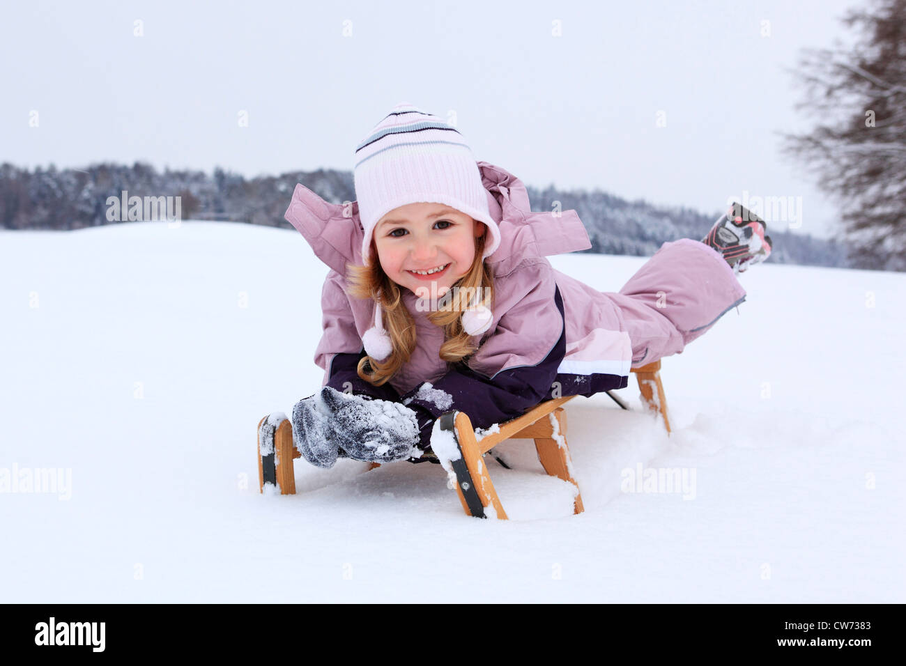 little girl in a snow field lying on the belly on a wooden toboggan ...