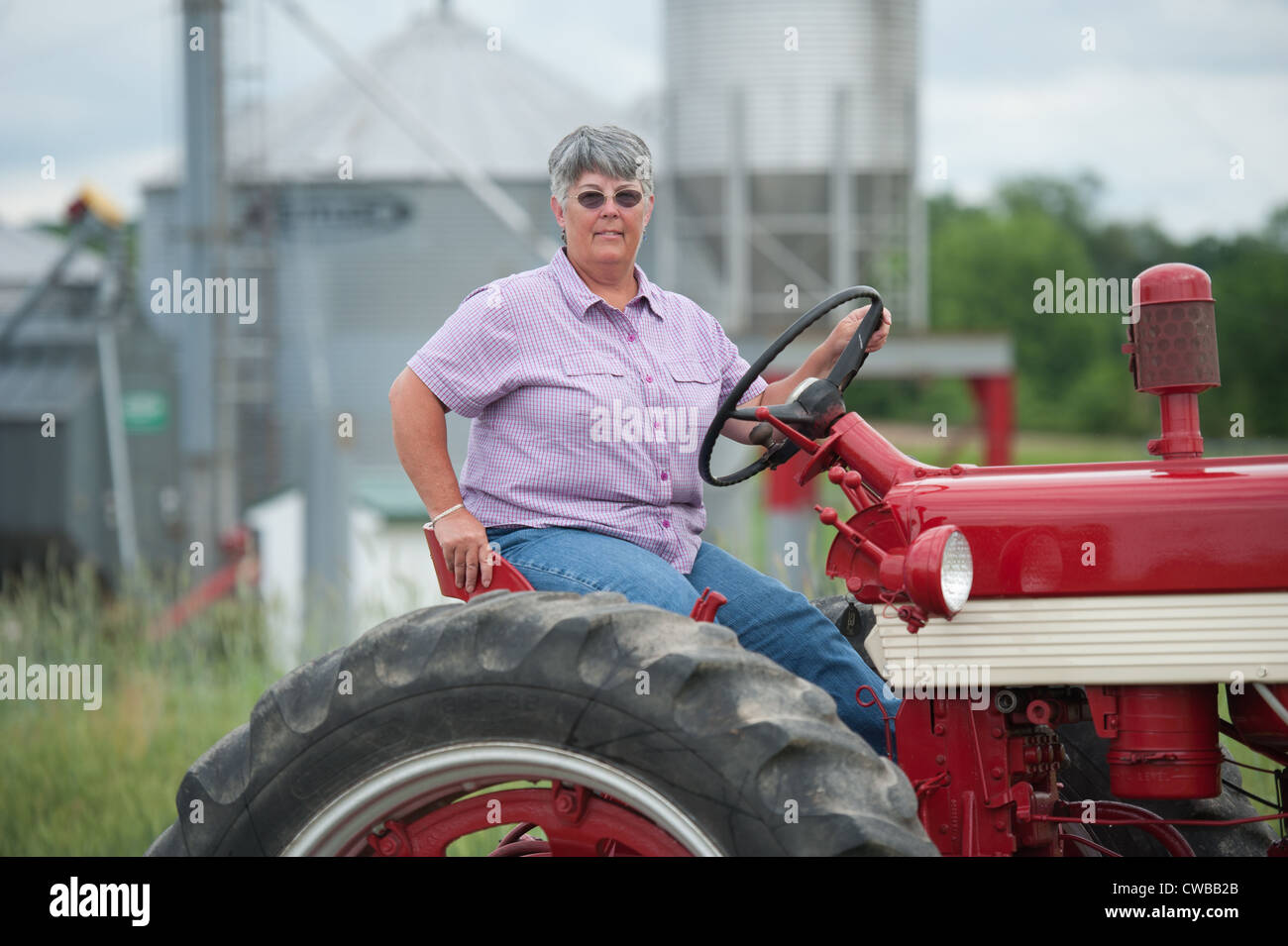 Woman farmer riding tractor in front of farm land in front of barns and ...
