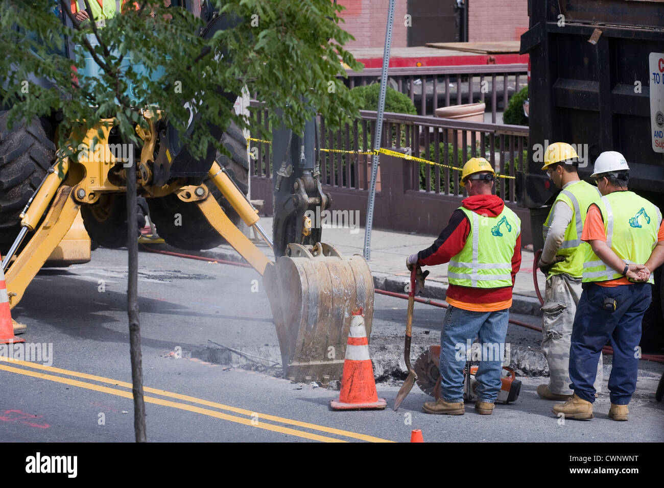Three Construction Workers watch a Backhoe excavate a hole in a roadway ...