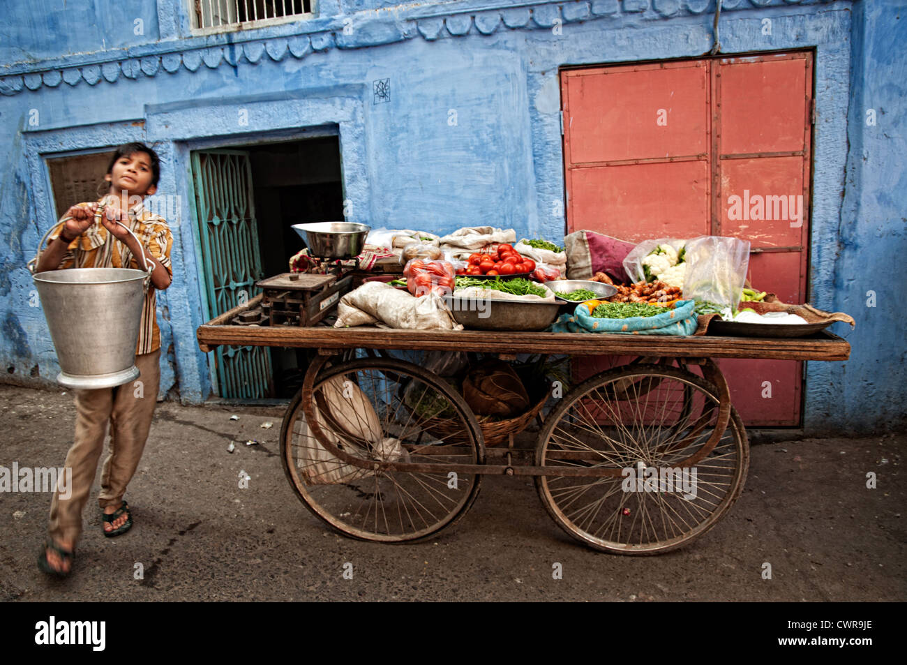 Boy carrying a bucket of water in front of a vegetables street stall ...