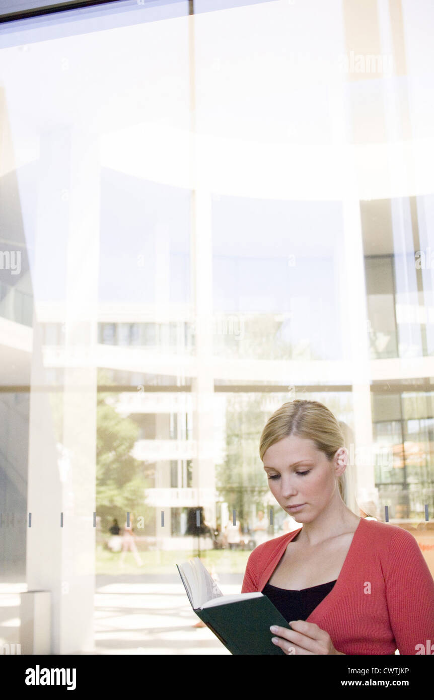Young woman leaning against glass wall reading book Stock Photo - Alamy