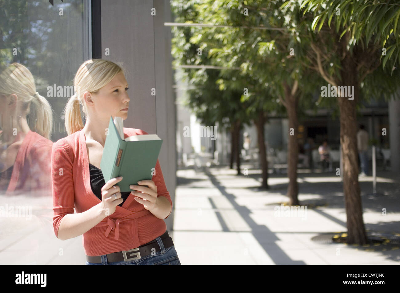 Young woman leaning against glass wall reading book Stock Photo - Alamy