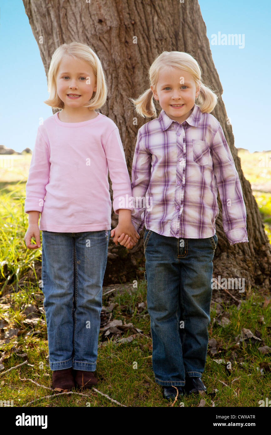 Twin blond girls are standing beside each other in front of a tree ...