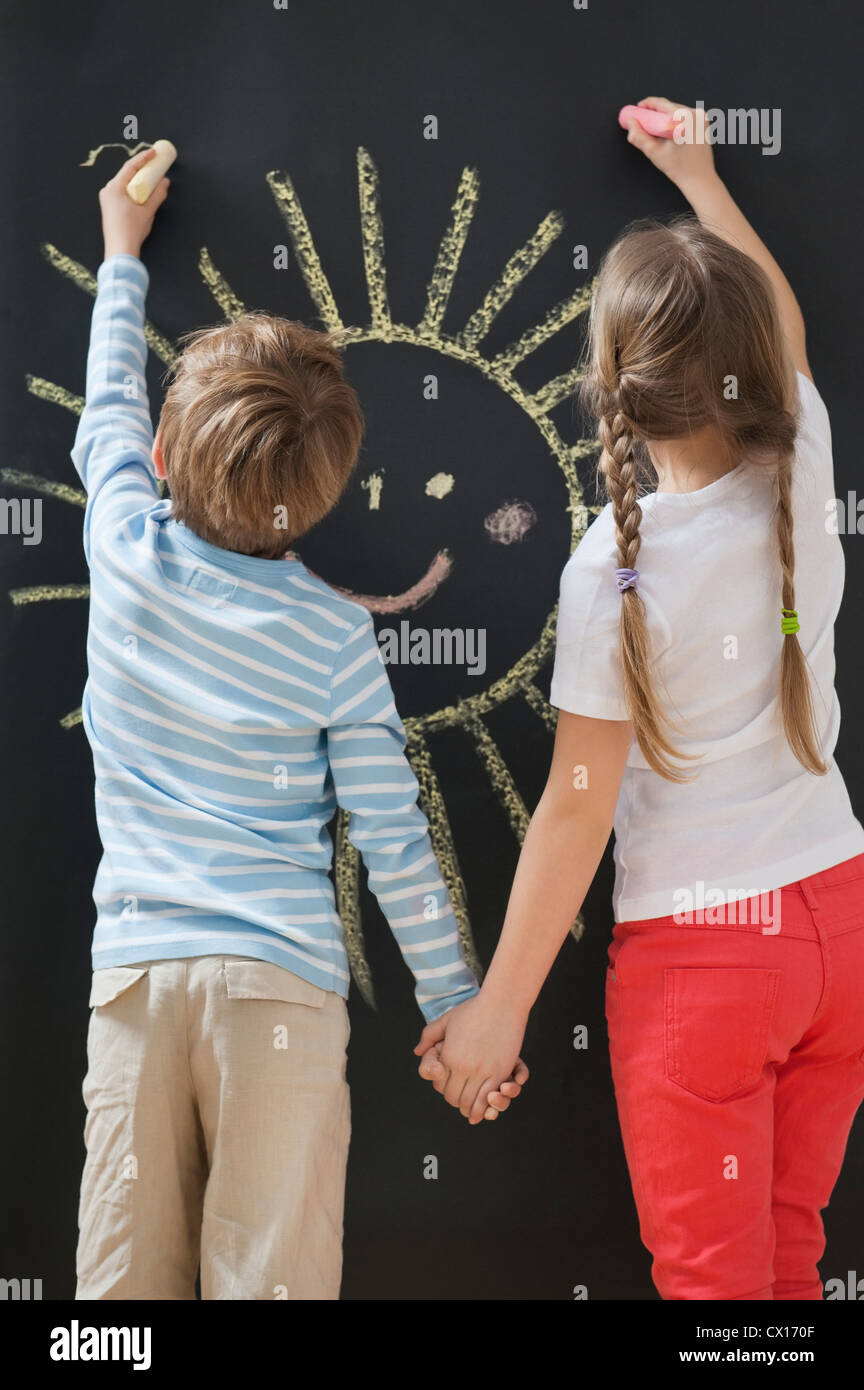 Back view of siblings drawing sun on blackboard while holding hands ...