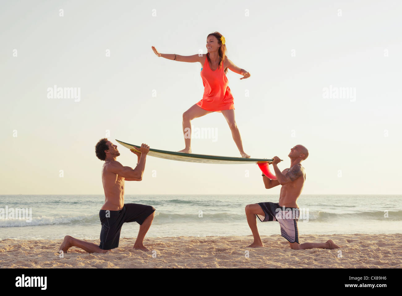 Two Men Hold A Surfboard As A Woman Stands On It In A Surfer Pose ...
