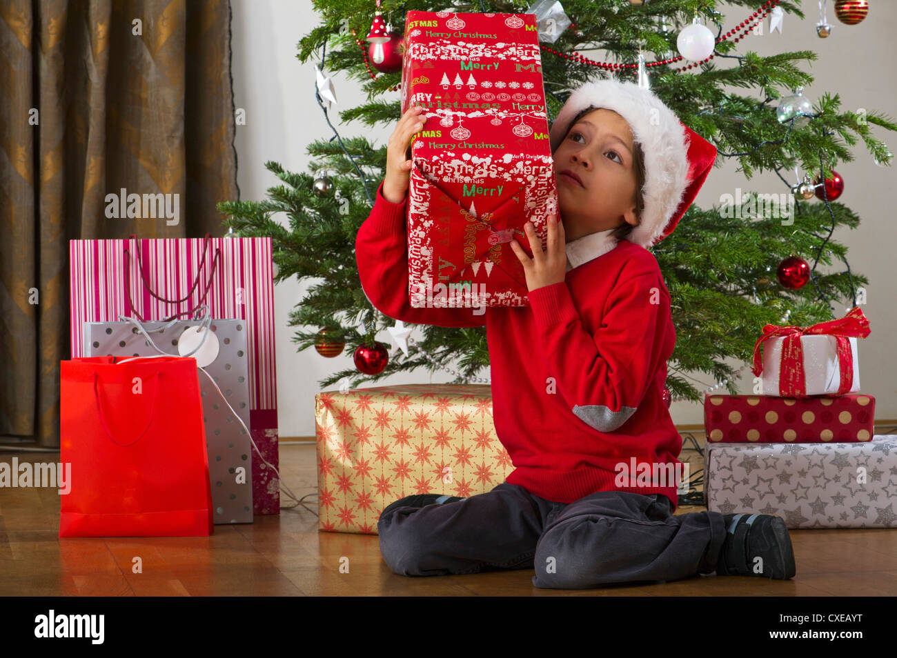 Boy sitting beside Christmas tree, shaking gift Stock Photo - Alamy