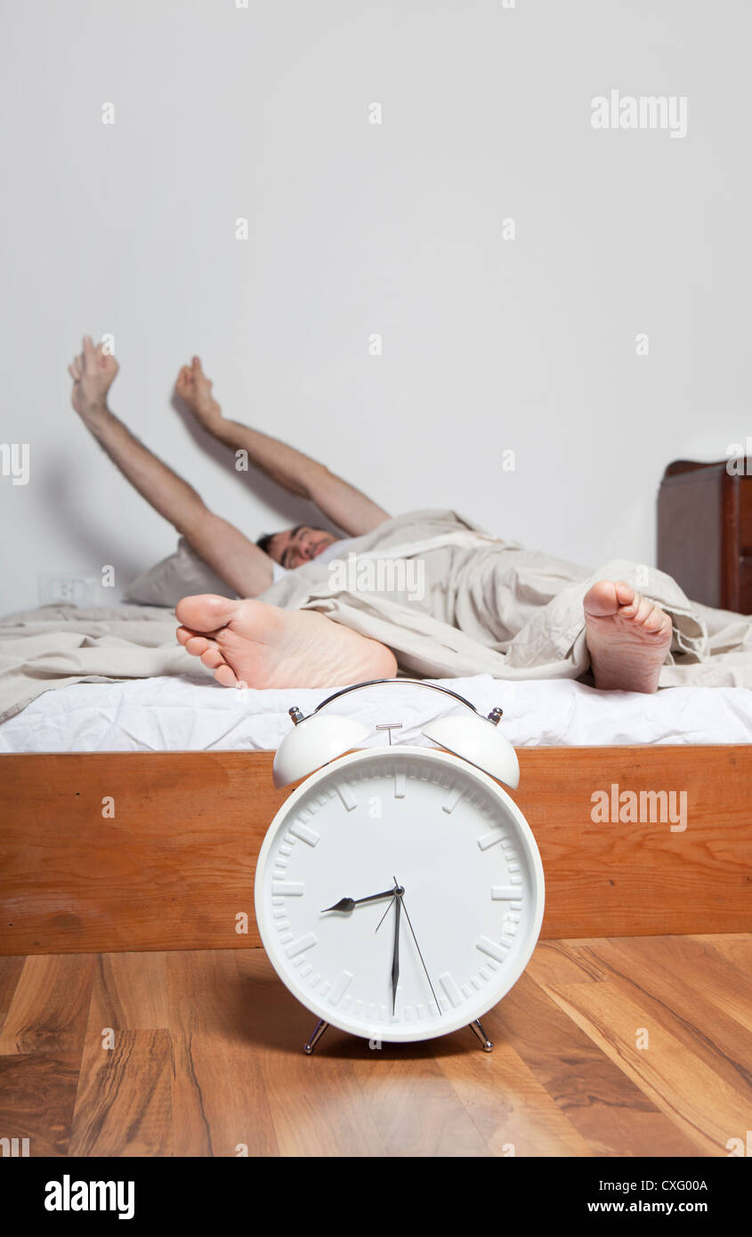 A young tired man laying on a bed with his arms raised. Big alarm clock ...