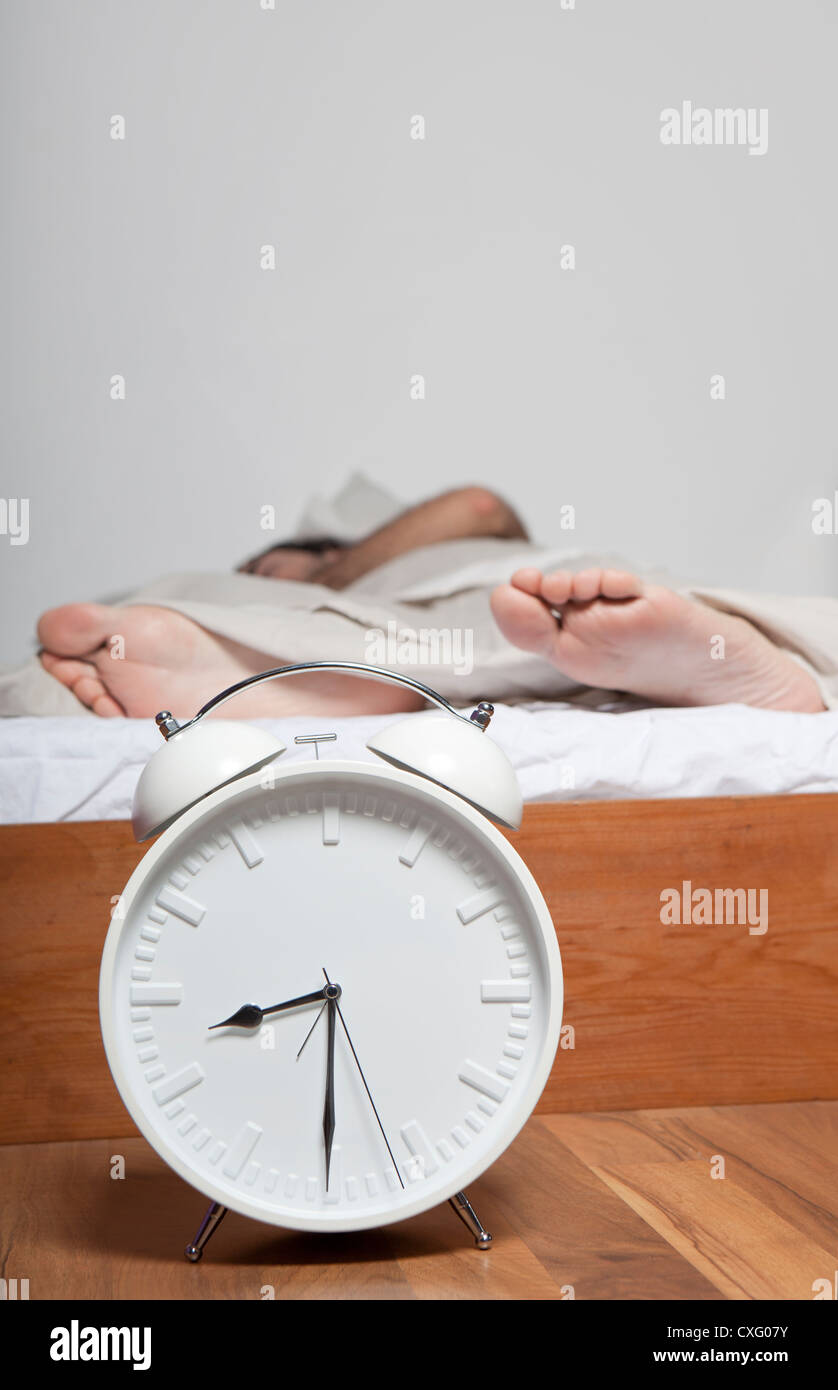 A young tired man laying on a bed. Big alarm clock is placed in front ...
