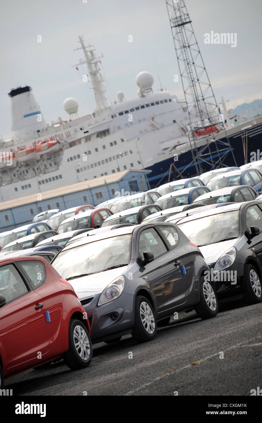 A row of newly imported cars in dock with passenger ship in background ...