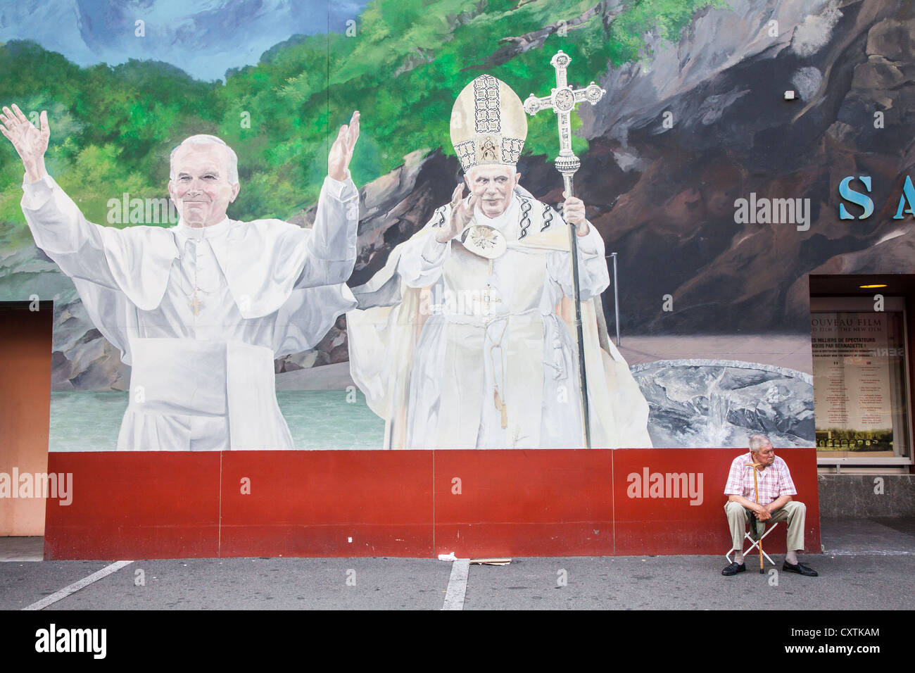 Man sitting on stool under a mural of the Pope in Lourdes, Hautes ...