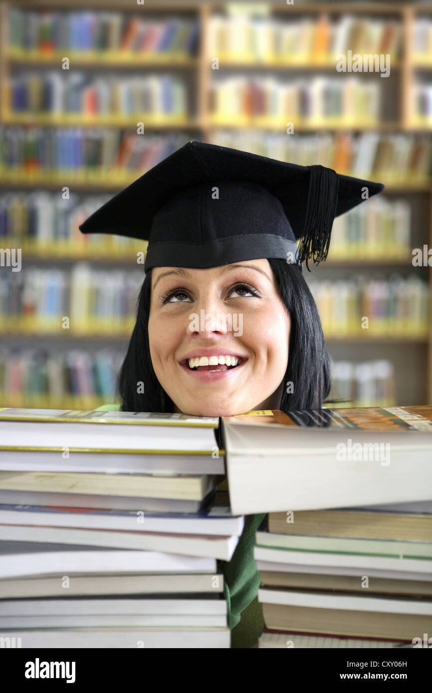 Smiling female student wearing a graduation cap behind stacks of books ...
