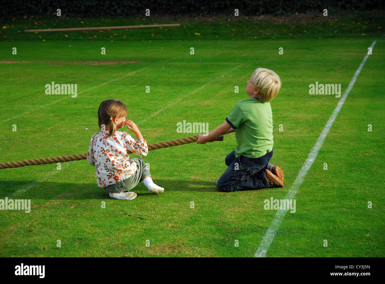 A young boy and girl play with a rope on a grass lawn. Their parents ...