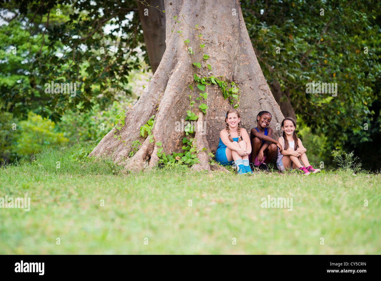 three 8-9 years old girls sitting near large tree and smiling at camera ...