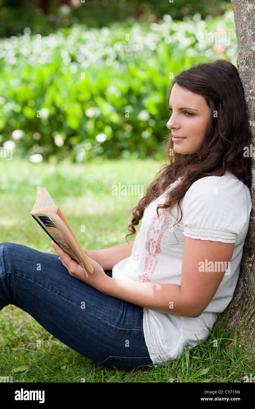 Young thoughtful woman leaning against a tree while reading a book ...