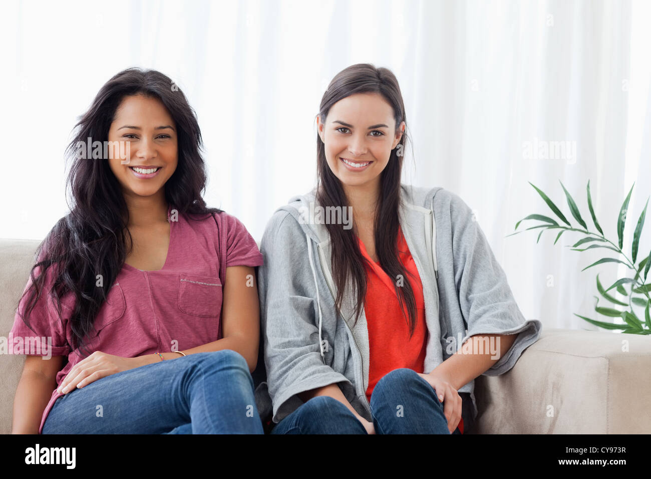 Two smiling women sit on the couch together while looking at the camera ...