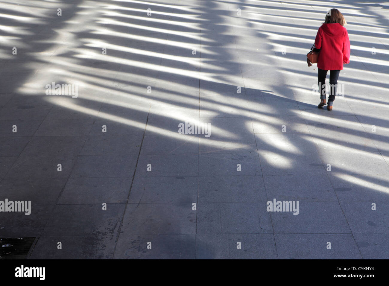 Woman in red coat walking away across vivid shadow on pavement, Madrid ...