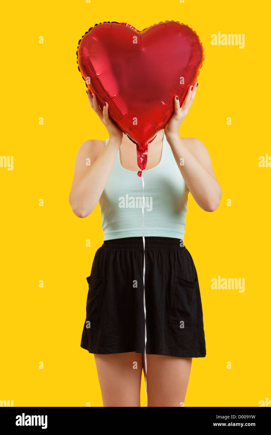 Young woman hiding behind heart shaped balloon over yellow background ...