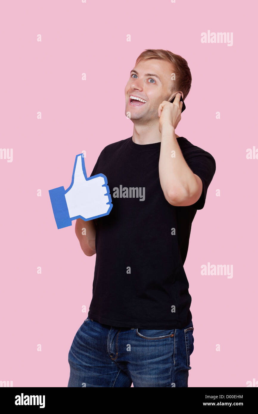 Cheerful young man using cell phone while holding fake like button over ...