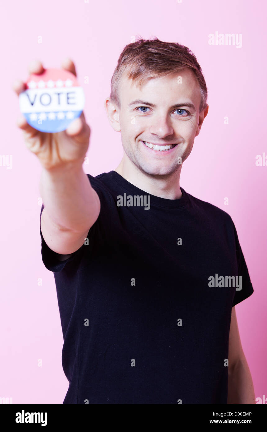 Portrait of a young man holding a VOTE badge up to the camera against ...