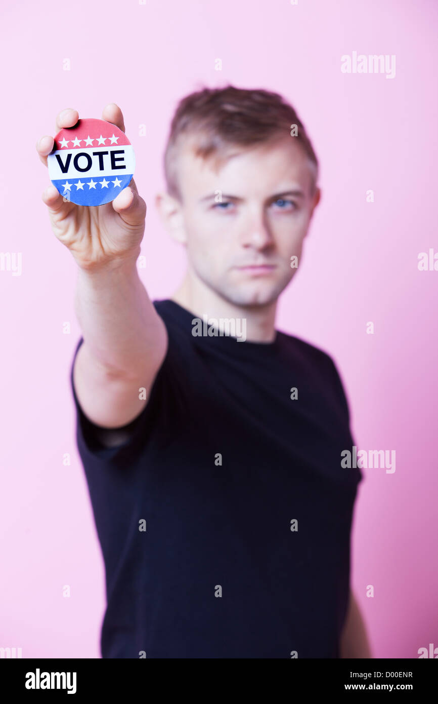 Portrait of a young man holding a VOTE badge up to the camera against ...