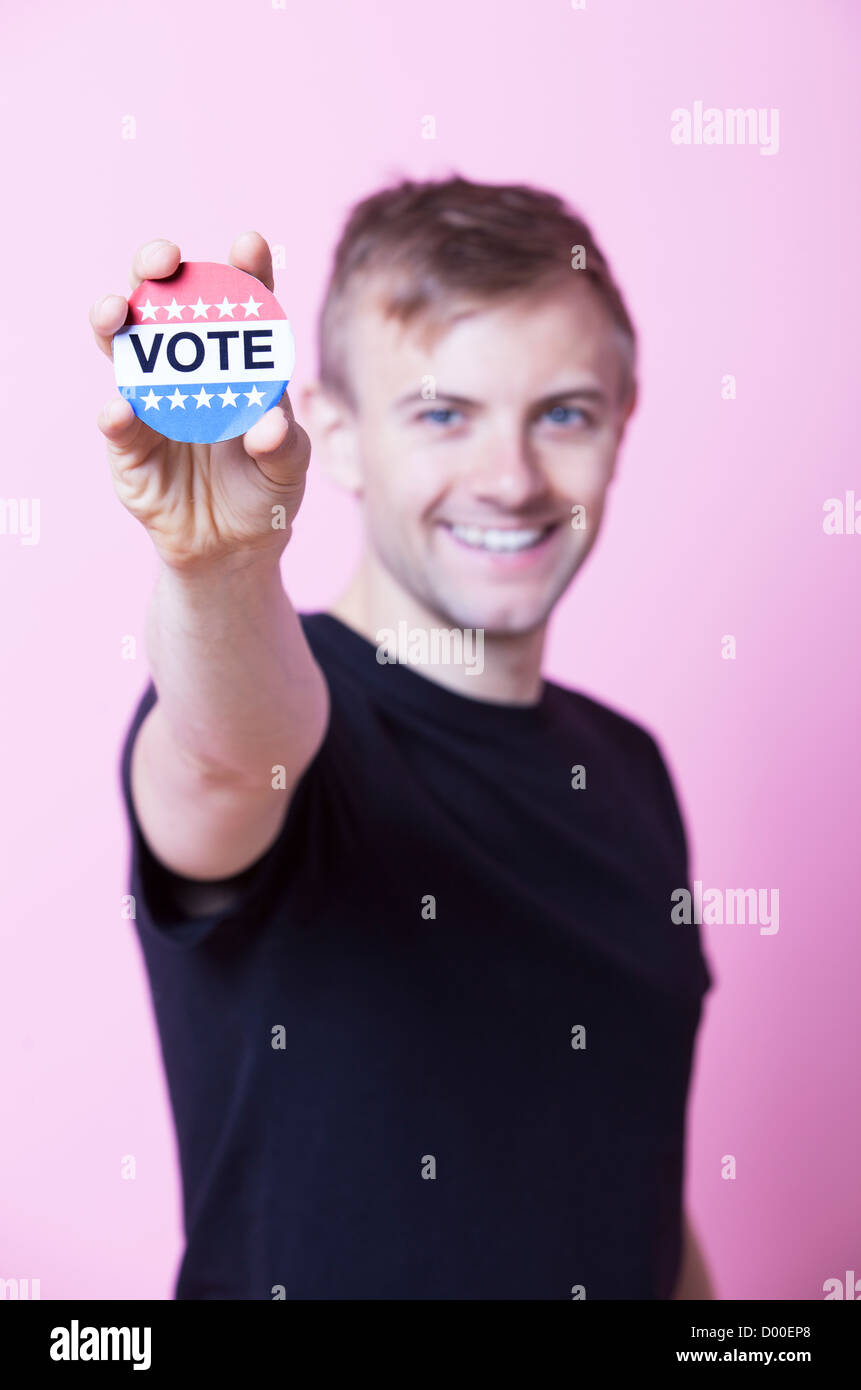 Portrait of a young man holding a VOTE badge up to the camera against ...