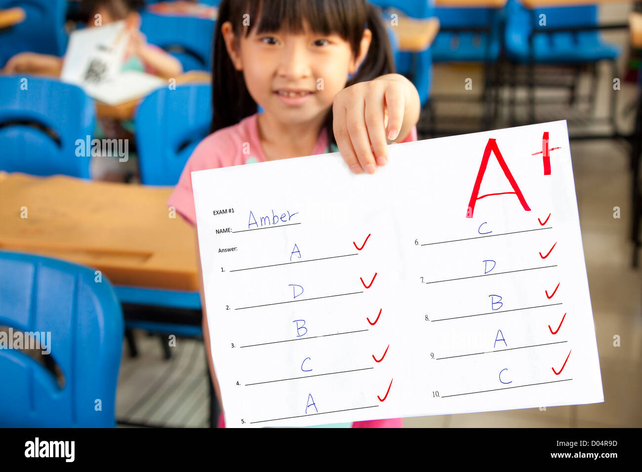 smiling little girl showing exam paper with a plus in the classroom ...