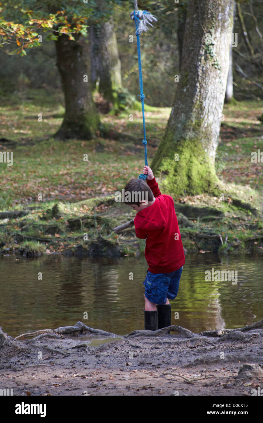 Young boy holding on to rope hanging on tree to swing over stream at at ...