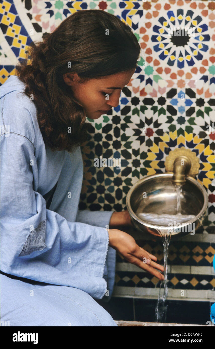 YOUNG MOROCCAN WOMAN IN A DJELLABA GETTING WATER IN A TASSA FROM THE ...