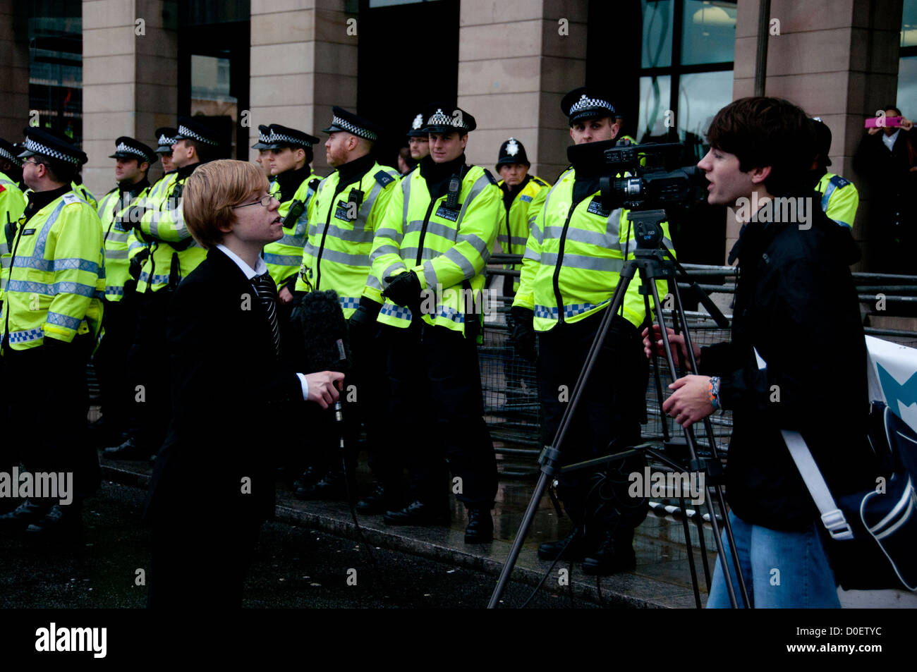 A young student reporter speaks to camera in front of a line of police ...