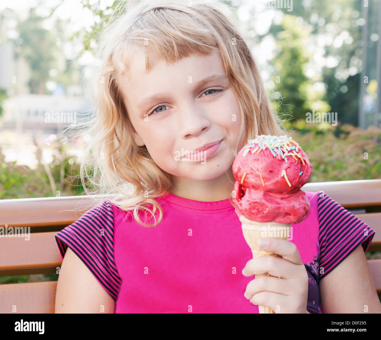 Little girl with big fruit ice-cream seats on wooden bench in park ...