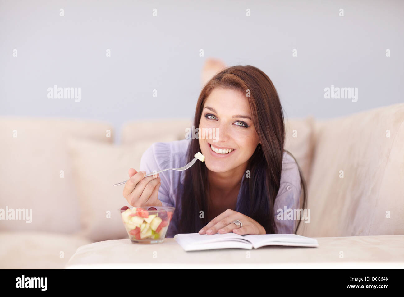 An attractive young woman eating fruit salad and reading while lying on ...