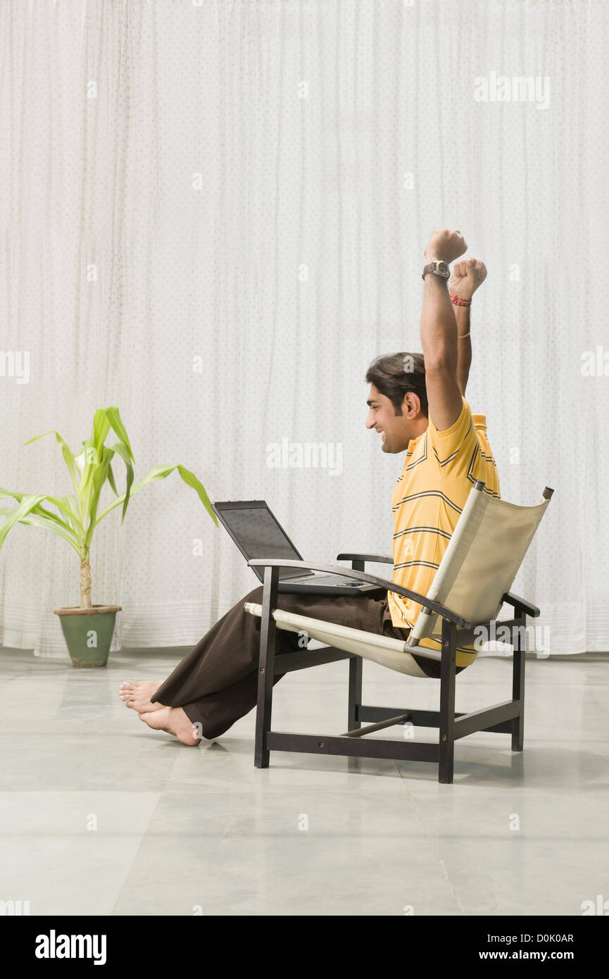 Young man sitting in an armchair with a laptop and his arms raised ...
