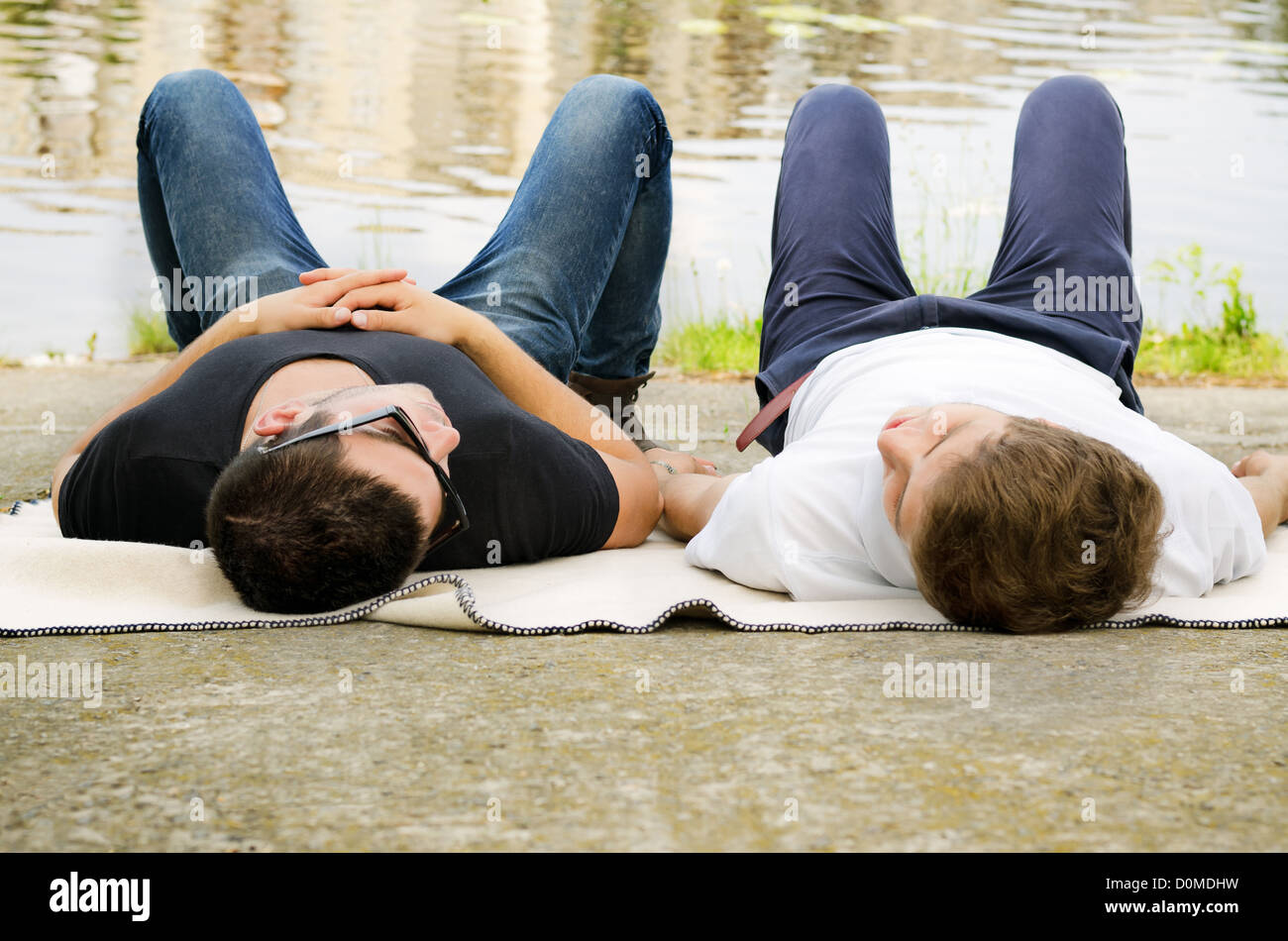 Two guys relaxing together lying in their jeans on a blanket alongside ...
