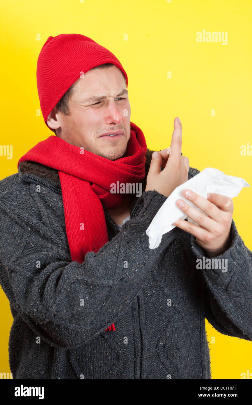 Ill young man with red nose, scarf and cap picking nose with hanky ...