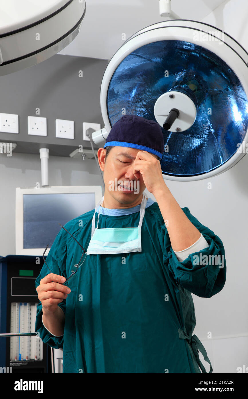 Doctor wearing scrubs standing in operating room rubbing his forehead ...
