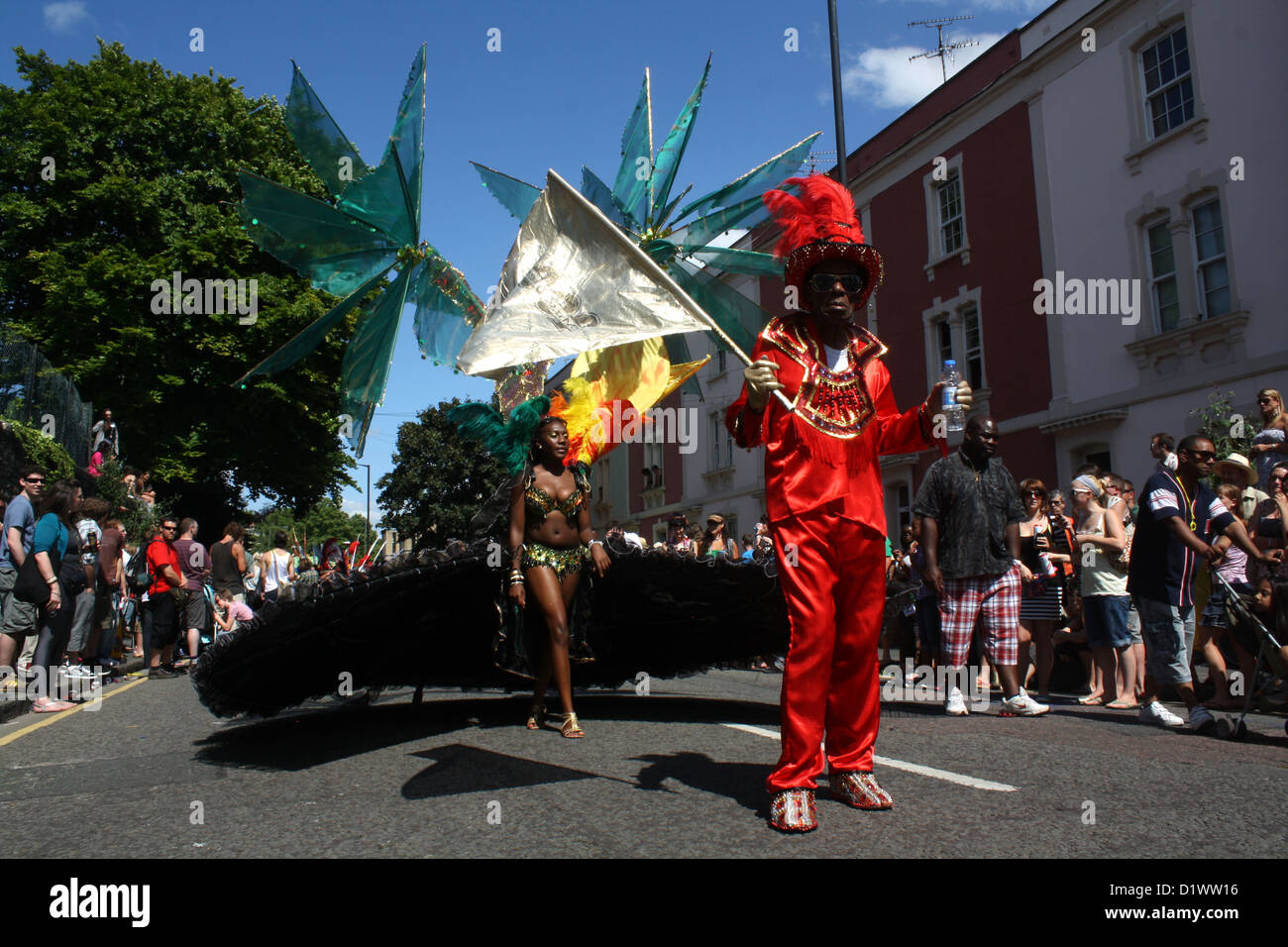 Carnival performers parade down a sun-lit street. A man in a red top ...