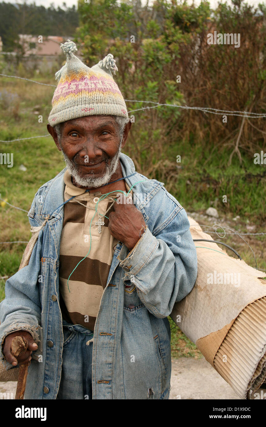 A homeless Hispanic man with a knit hat carrying a cane and a cardboard ...