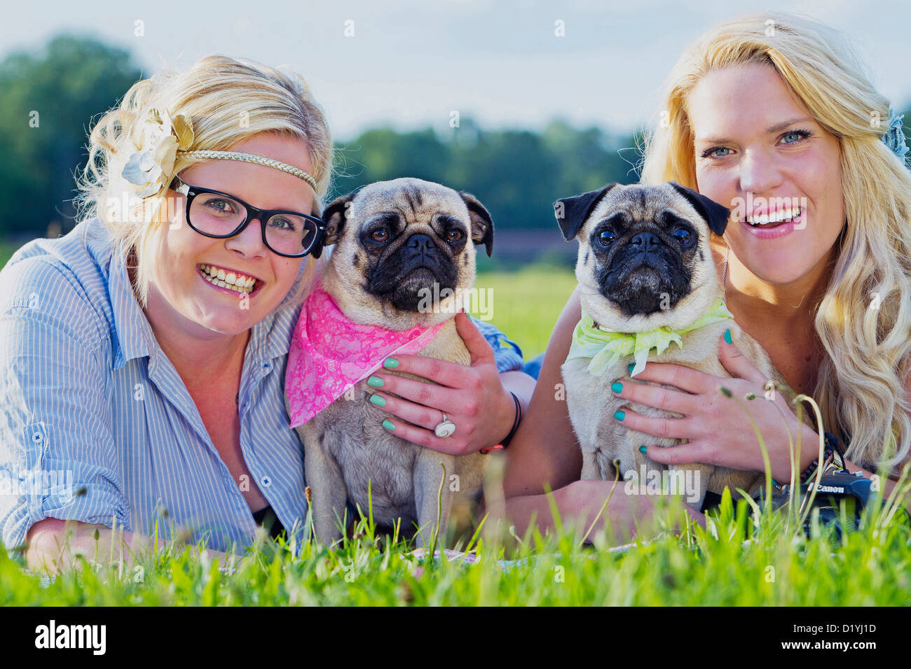 Domestic Dog, Pug. Two smiling, blonde women lying on a meadow in ...