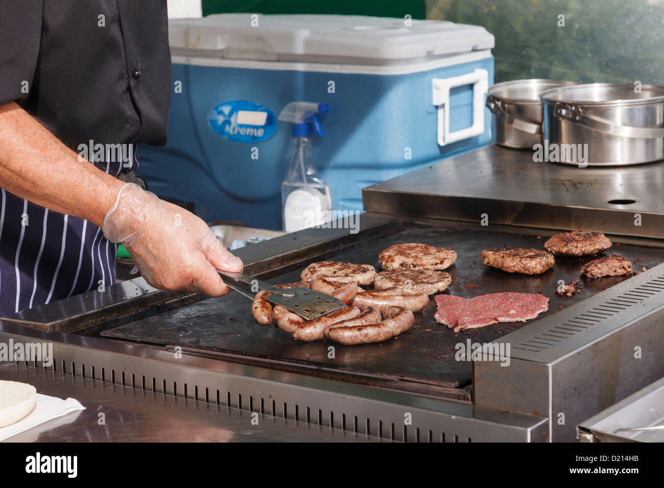 Man cooking sausages burgers and steak on a food stall griddle at ...