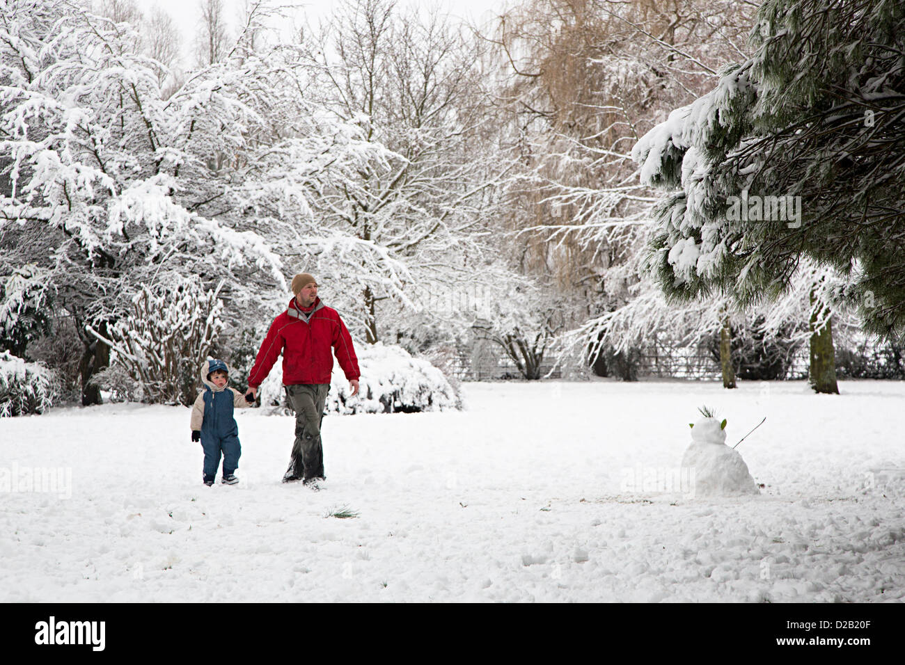 Man and child holding hands walking in snow in public park with snowman ...