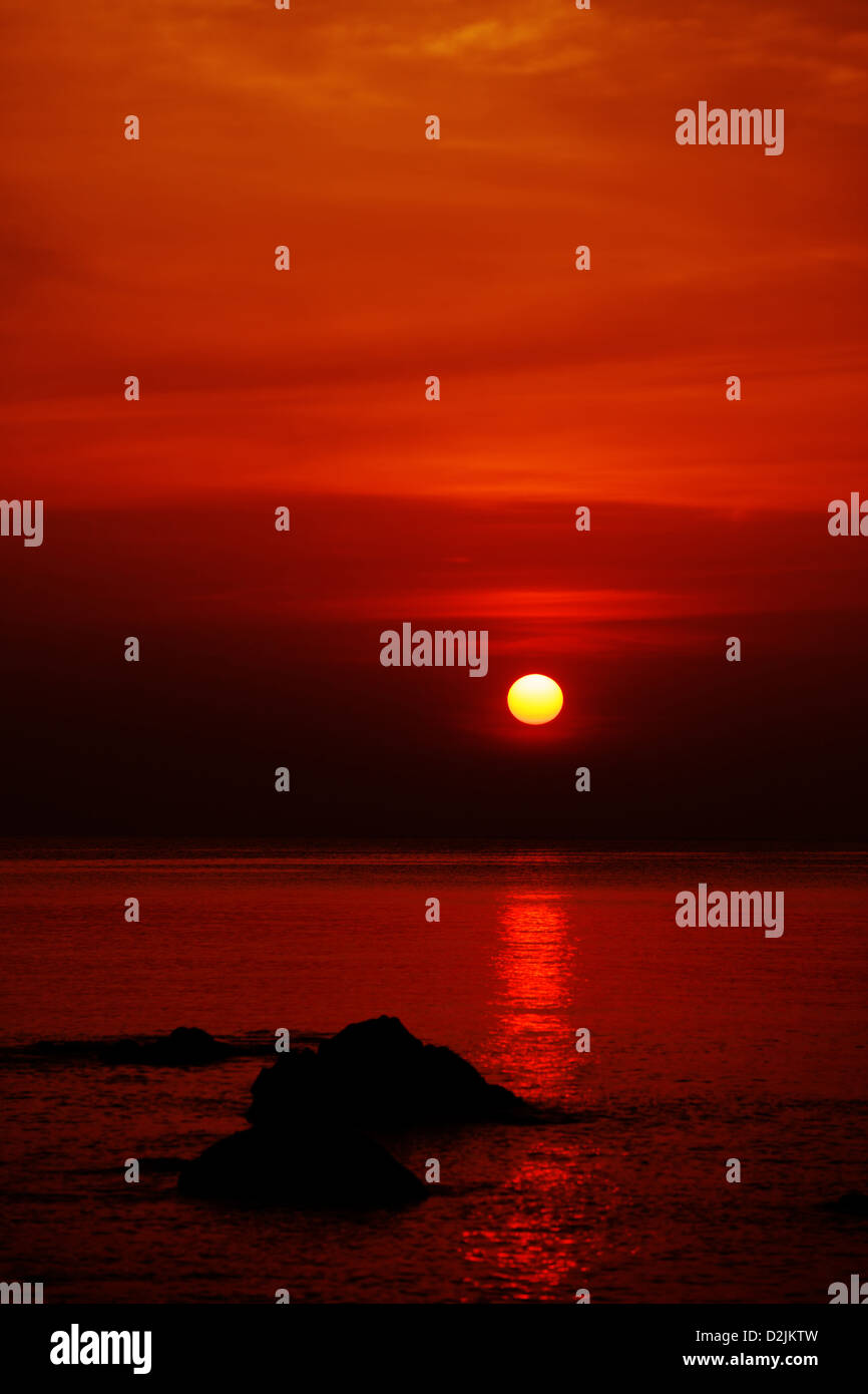Bright red colored sunset under calm smooth sea with stone on front ...