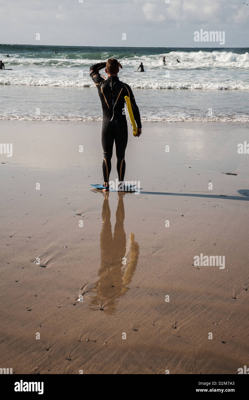 Surfer stands on the beach and looks out to sea at the surf in Newquay ...