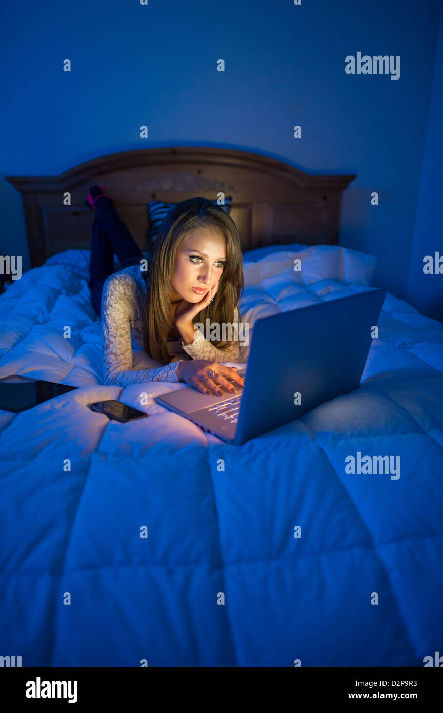 Beautiful teenage girl lying on a bed at night working on a laptop ...