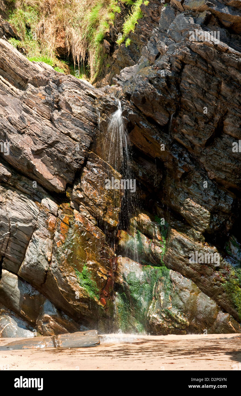 A small trickle of water flows over the rock face onto a sandy beach ...