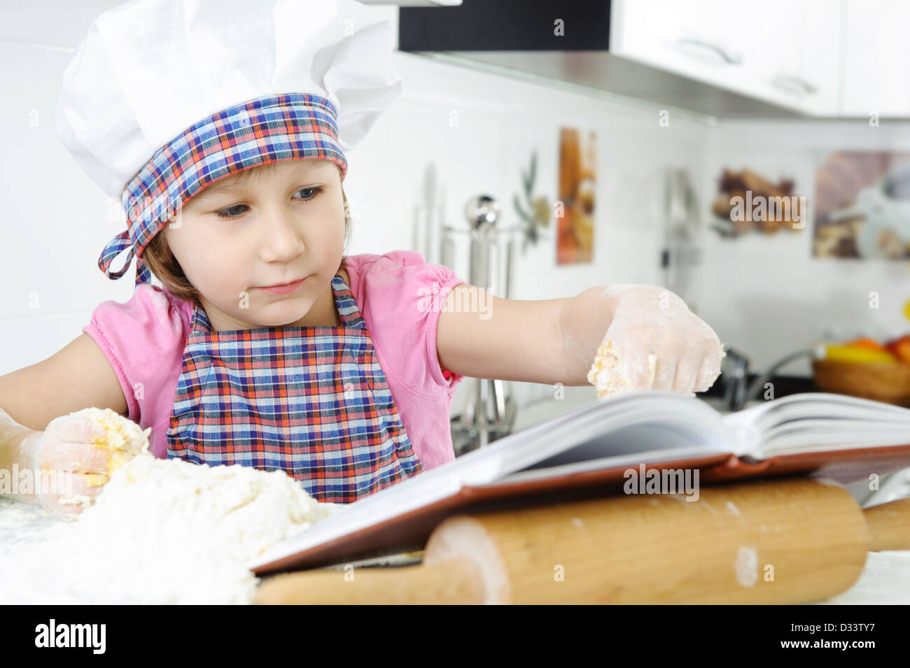 Little girl in hat and apron preparing cookies with cookbook Stock ...