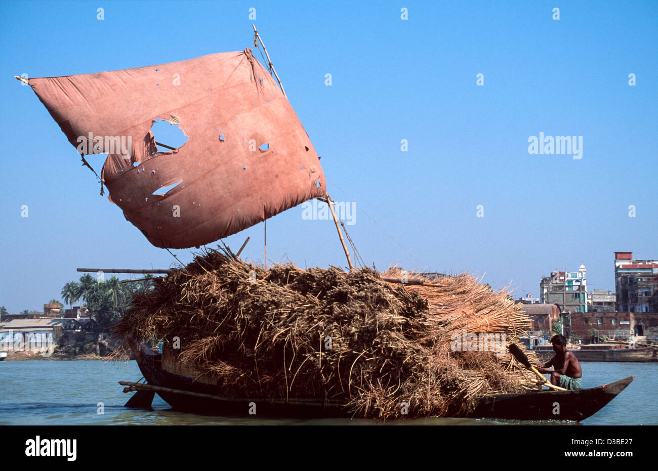 Small motorised boat under ragged sail and laden with bundles of jute ...