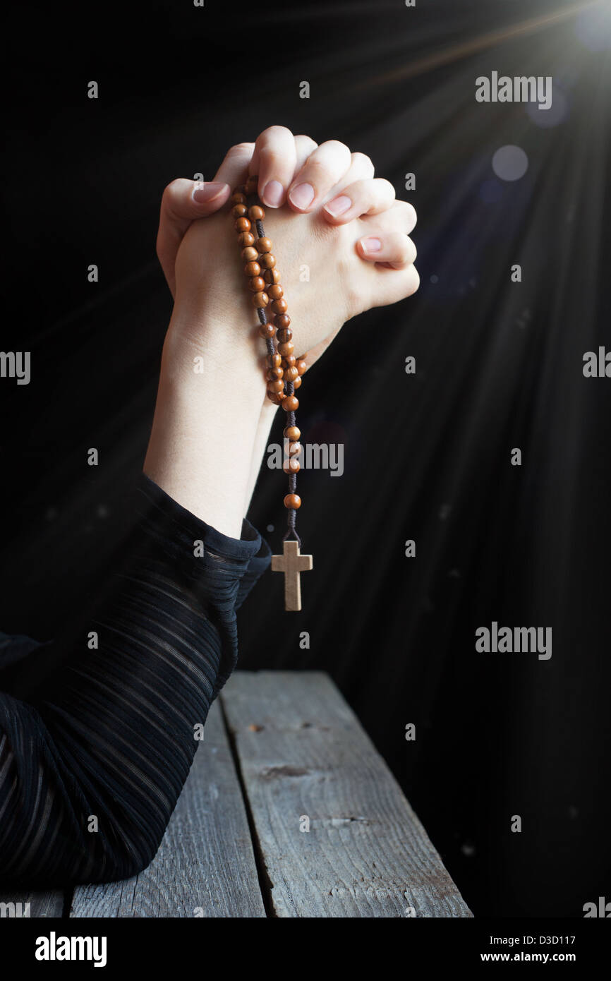Hands holding rosary with a cross and folded in prayer in the rays of ...