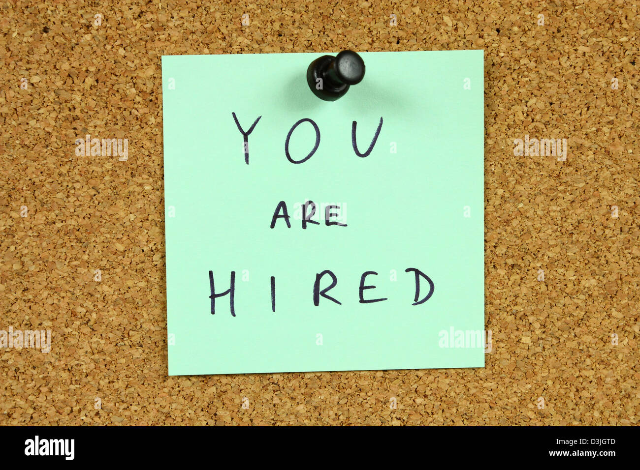 Green small sticky note on an office cork bulletin board. You are hired ...